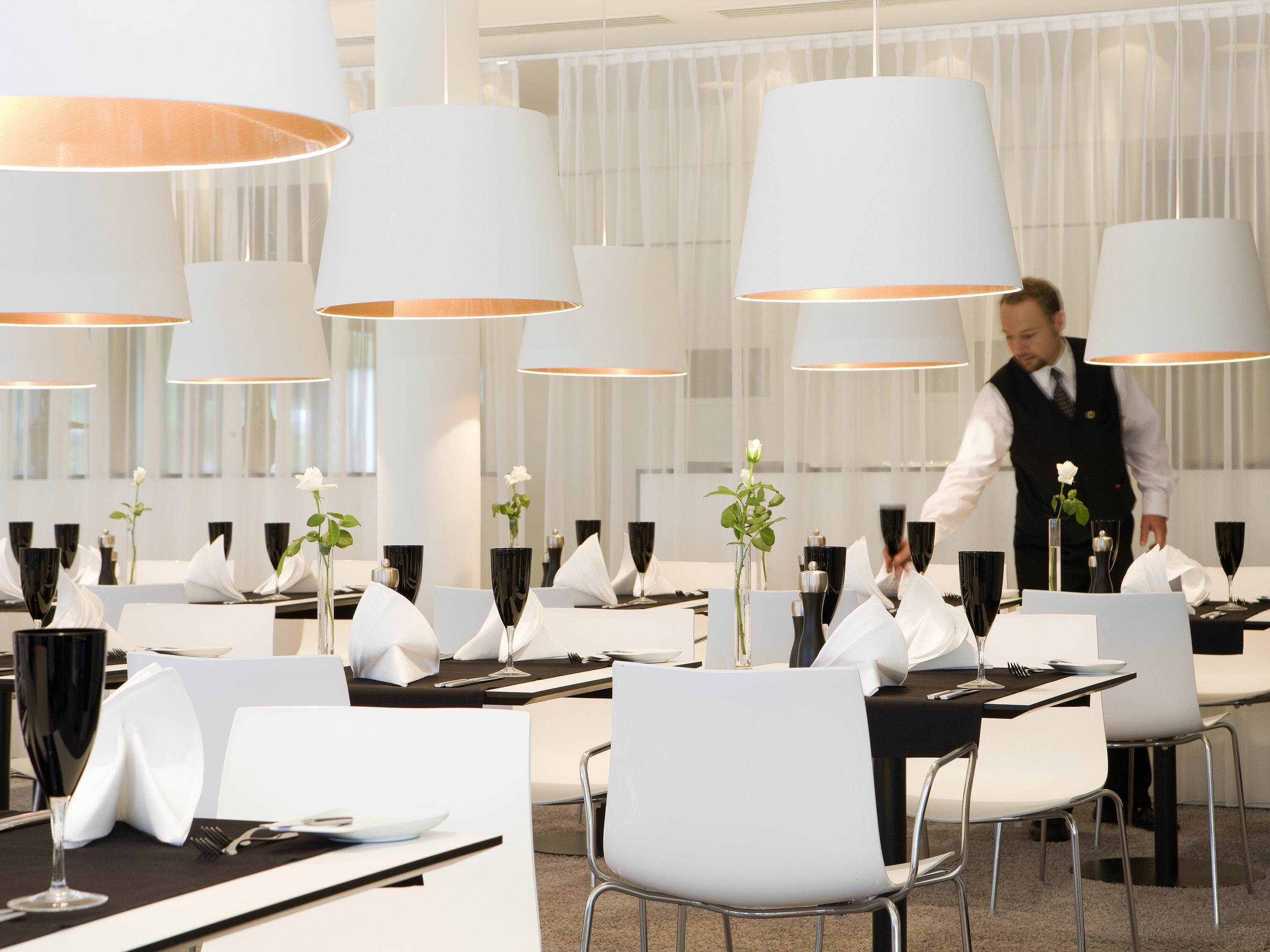 Uma sala de jantar moderna com mesas brancas, cadeiras pretas e grandes luminárias pendentes.