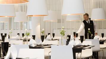 Uma sala de jantar moderna com mesas brancas, cadeiras pretas e grandes luminárias pendentes.