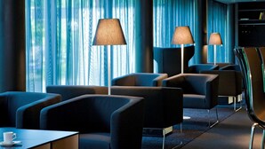 Quarto de hotel moderno com área de estar, janelas amplas com cortinas e duas luminárias de pé.