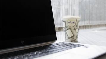 Um laptop sobre uma superfície branca, com uma xícara de café ao lado, identificada com a inscrição "nhow BRUSSELS".