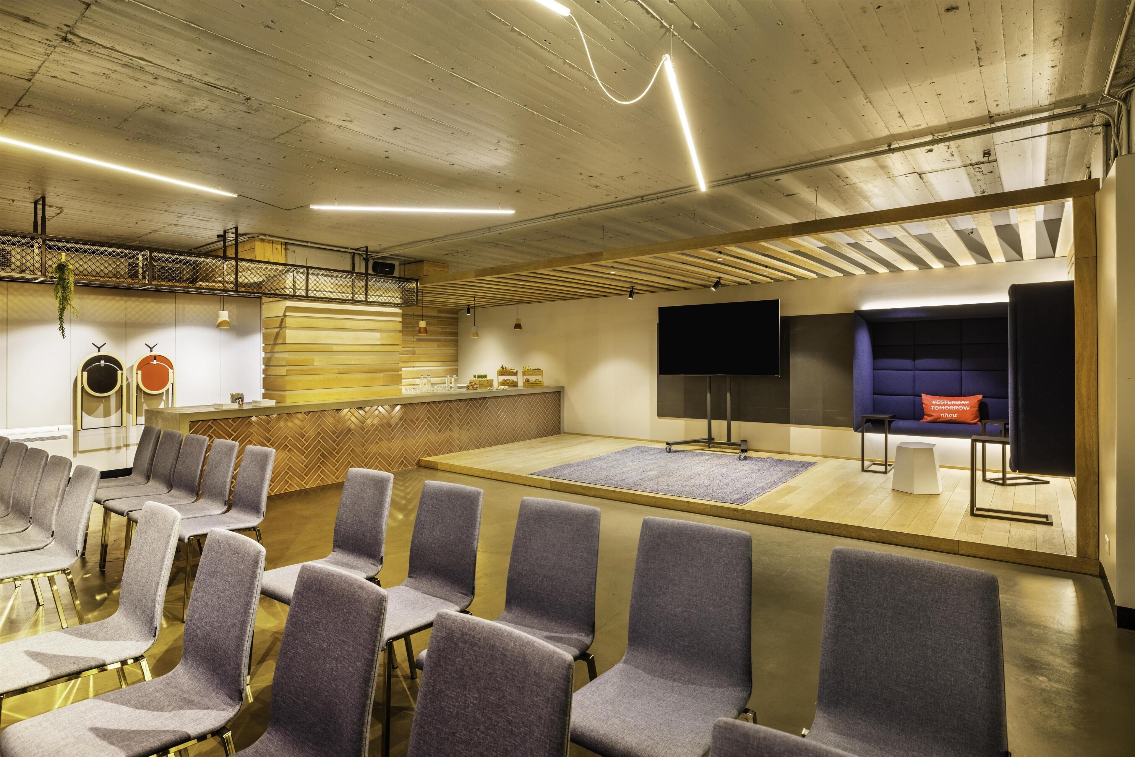Uma sala de conferências com uma mesa comprida, várias cadeiras, um palco e uma tela grande.