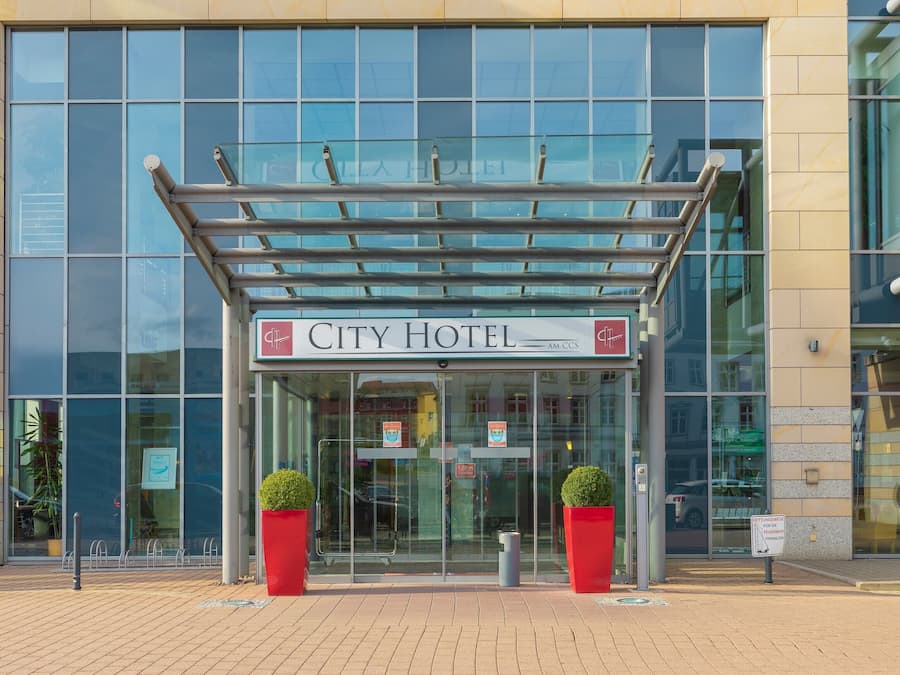 Entrada do The City Hotel com fachada de vidro, toldo metálico e vasos vermelhos.