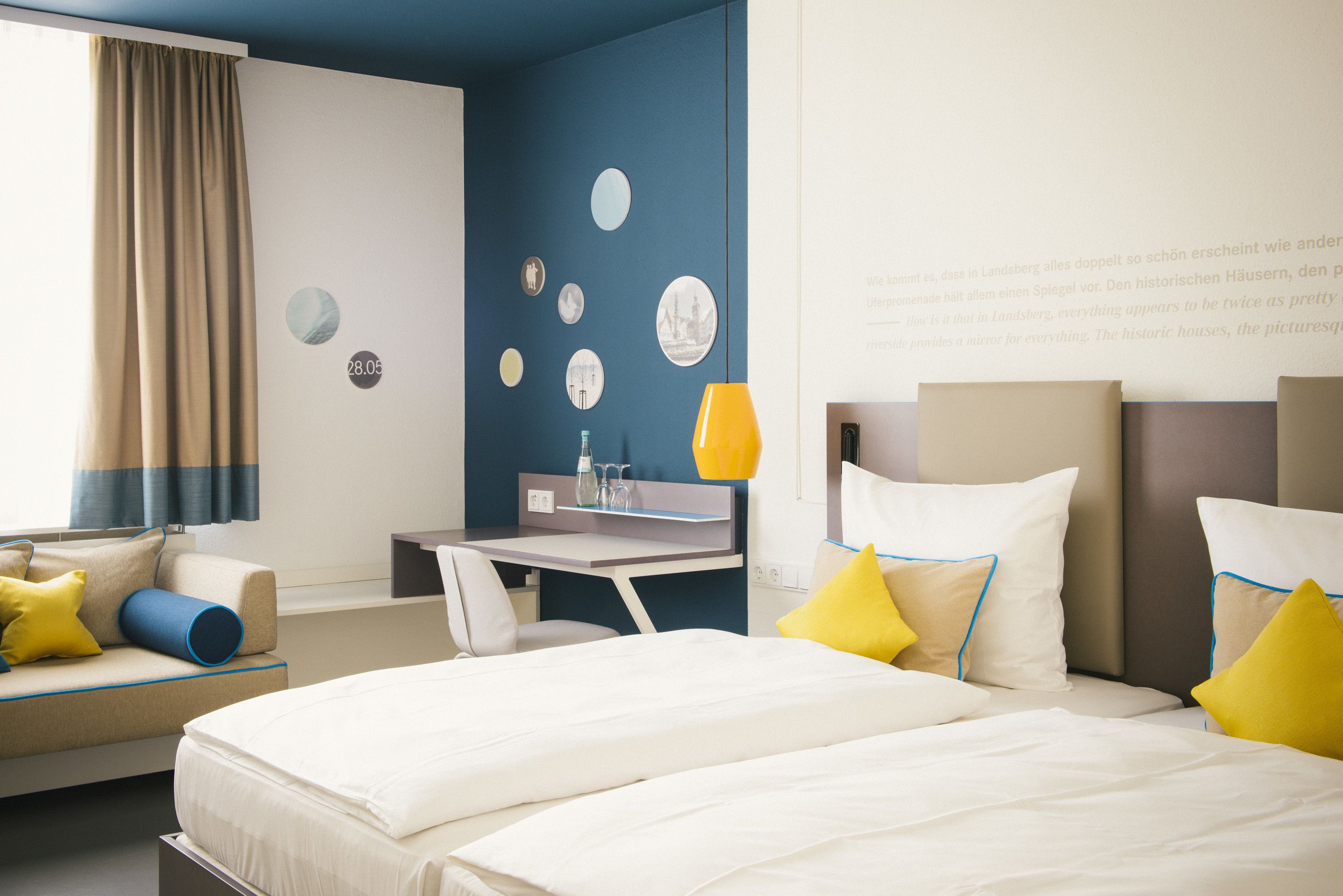 Quarto de hotel com cama, escrivaninha e cadeira. Há almofadas e uma luminária na cama.