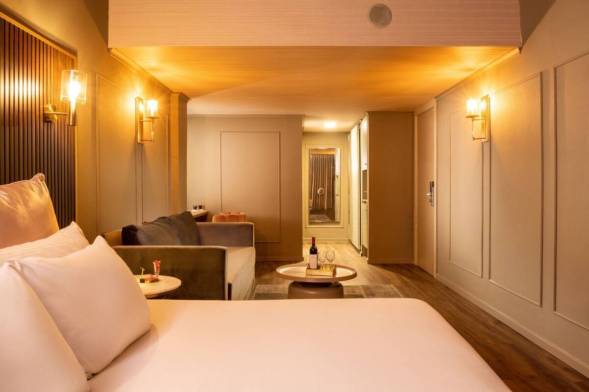 Quarto de hotel com cama, sofá, mesa pequena com garrafa e copos, e espelho.