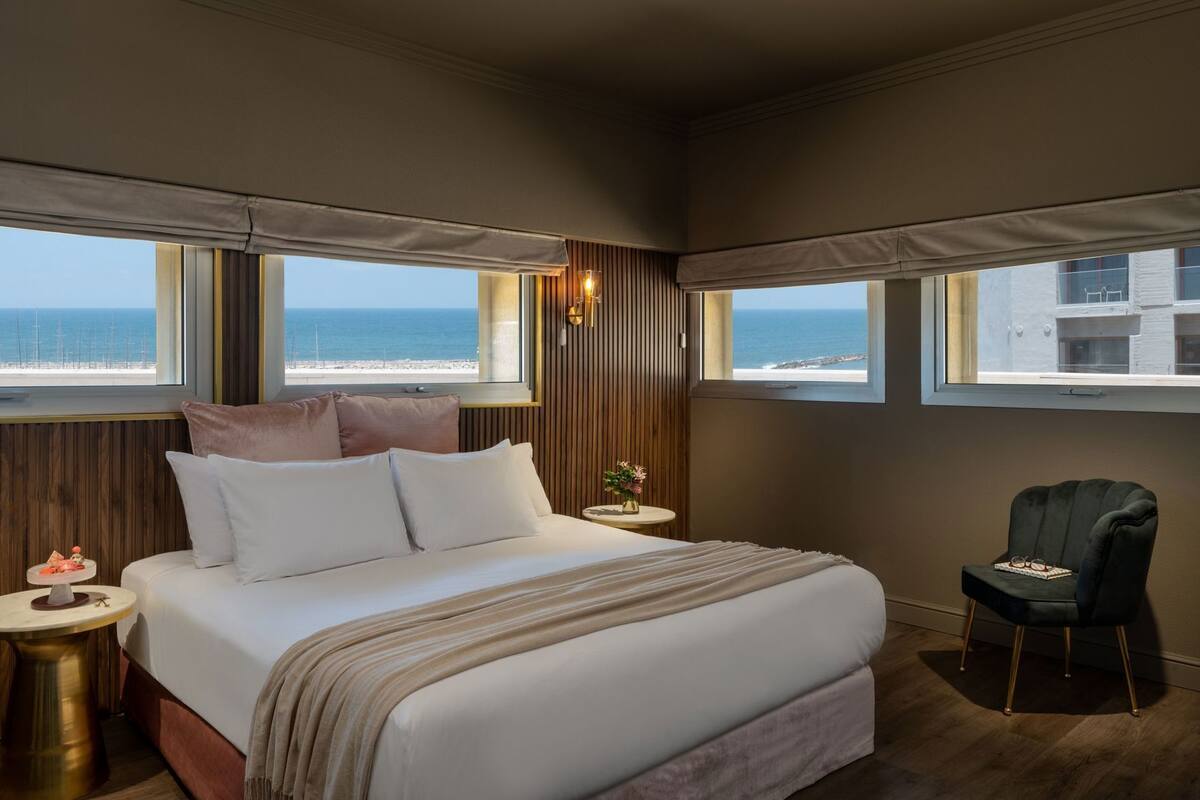 Quarto com cama grande, duas mesas de cabeceira, uma cadeira e vista para a praia.