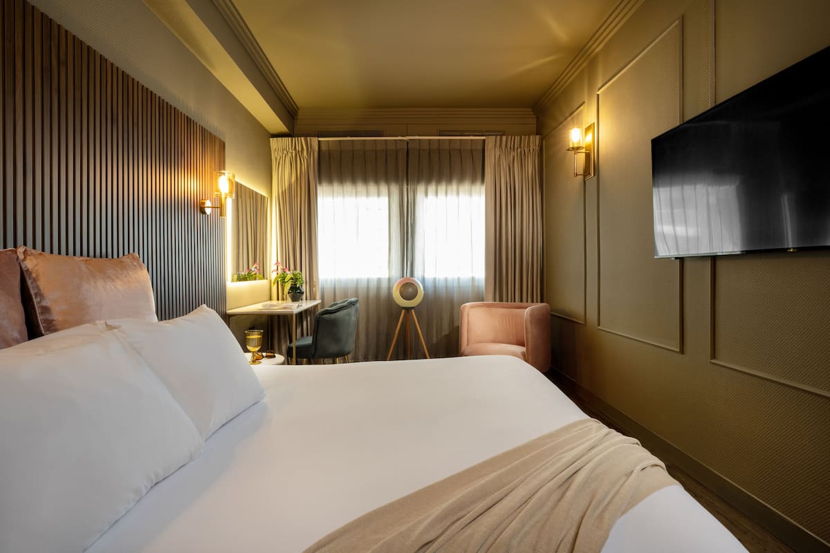 Quarto de hotel com uma cama grande, uma escrivaninha, uma cadeira, uma televisão e uma janela com cortinas.