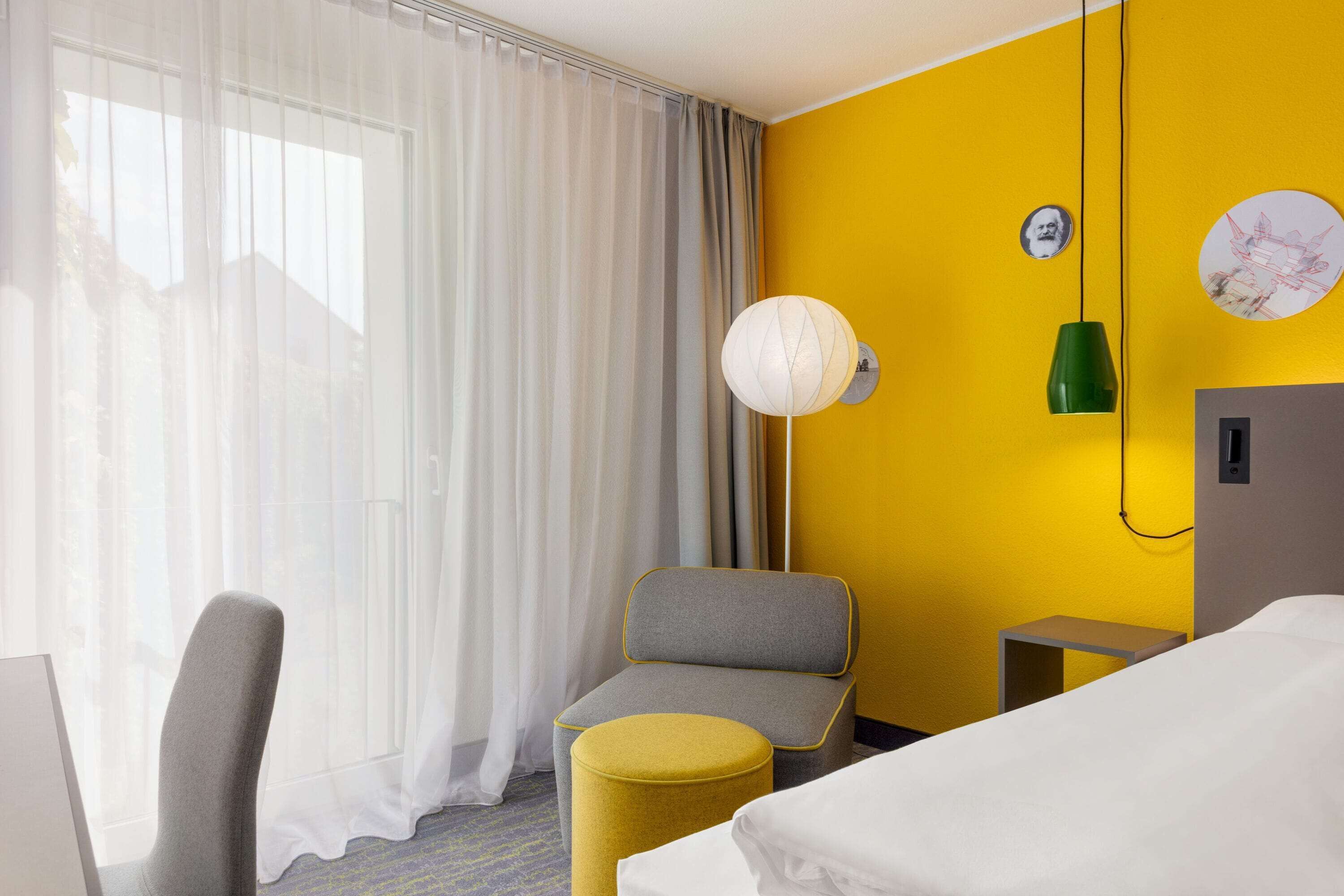 Quarto de hotel com cama, uma cadeira, mesinha, luminária e janela com cortinas.