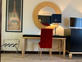 Quarto de hotel com escrivaninha, uma cadeira vermelha, um suporte preto para televisão, luminária e quadro com a Torre Eiffel.
