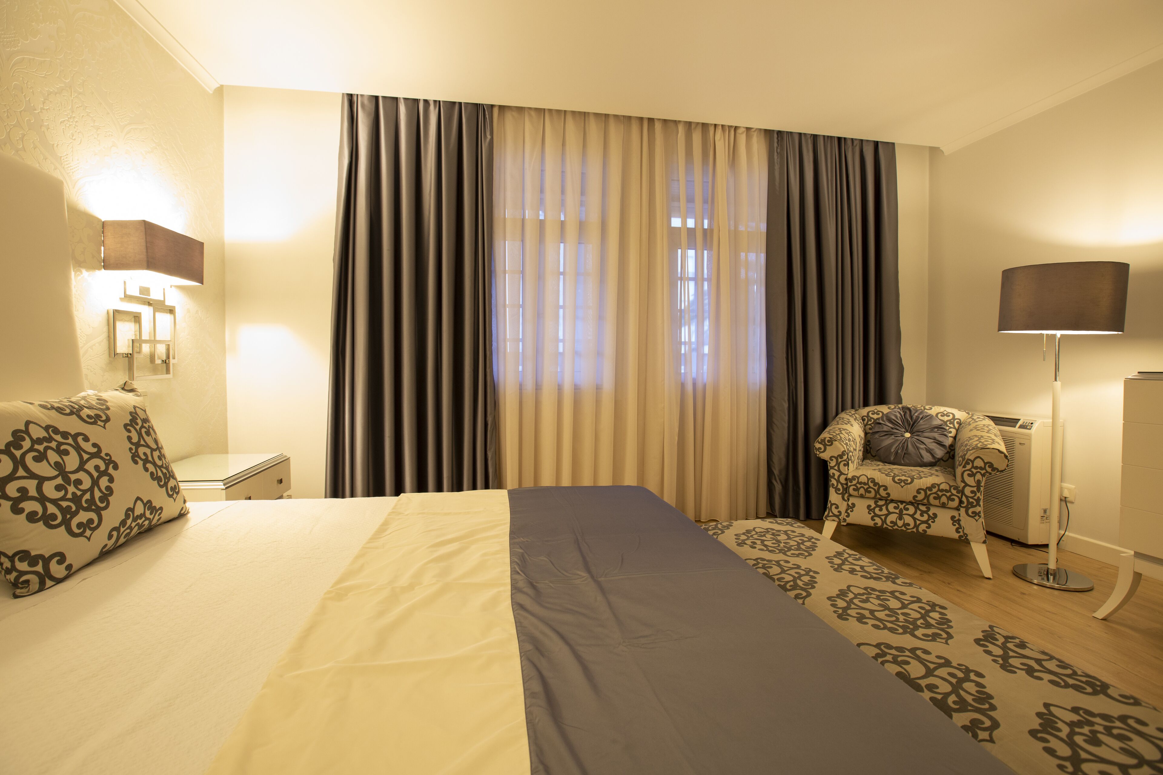 Quarto de hotel com cama, cadeira, abajur e janela com cortinas.