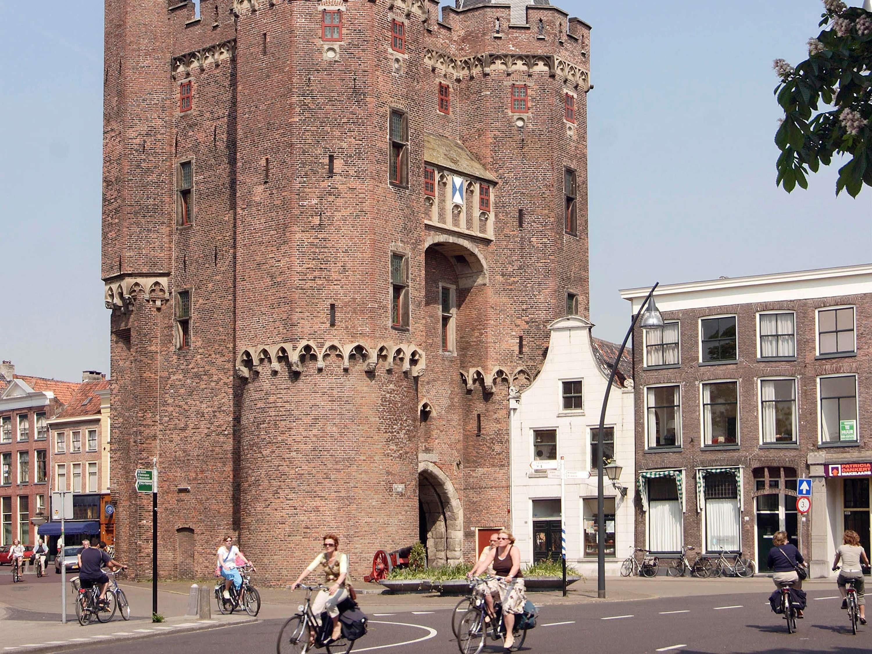 Uma torre de tijolos histórica com turreliha, rodeada por edifícios modernos e ciclistas na rua.