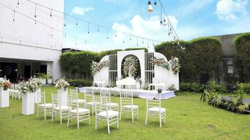 Montagem de casamento ao ar livre com cadeiras brancas dispostas em fileiras, palco com toldo branco e decorações florais.