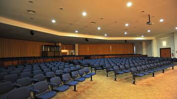Um amplo auditório com fileiras de cadeiras azuis, um palco com cortinas e piso coberto de carpete.