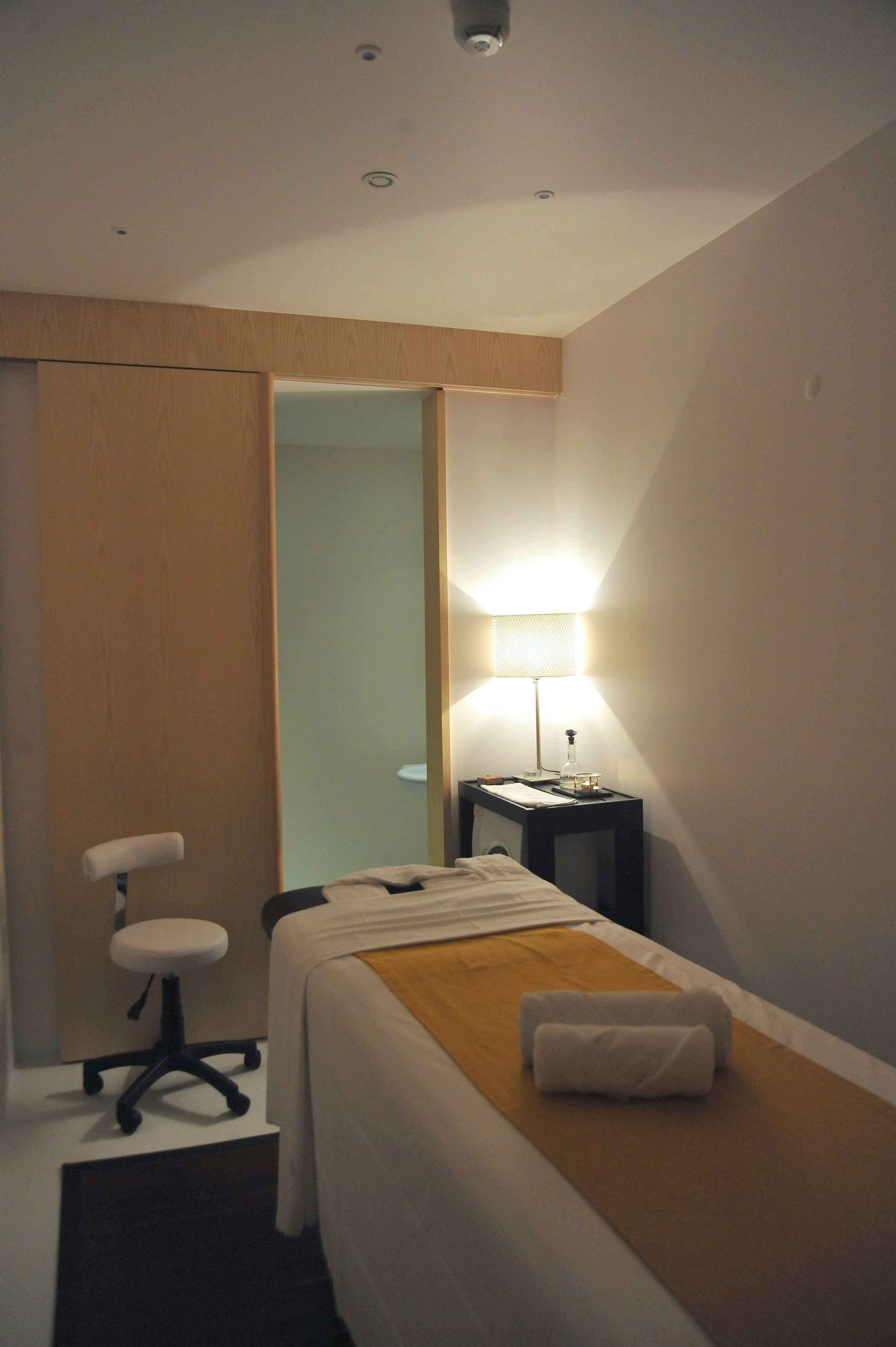 Quarto de hotel com uma cama de solteiro, escrivaninha, cadeira e mesa de cabeceira com abajur.