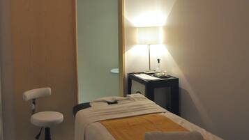 Quarto de hotel com uma cama de solteiro, escrivaninha, cadeira e mesa de cabeceira com abajur.