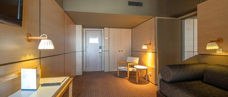 Quarto de hotel com cama, escrivaninha, cadeira e uma pequena mesa. Há uma televisão fixada na parede e uma janela com persianas.
