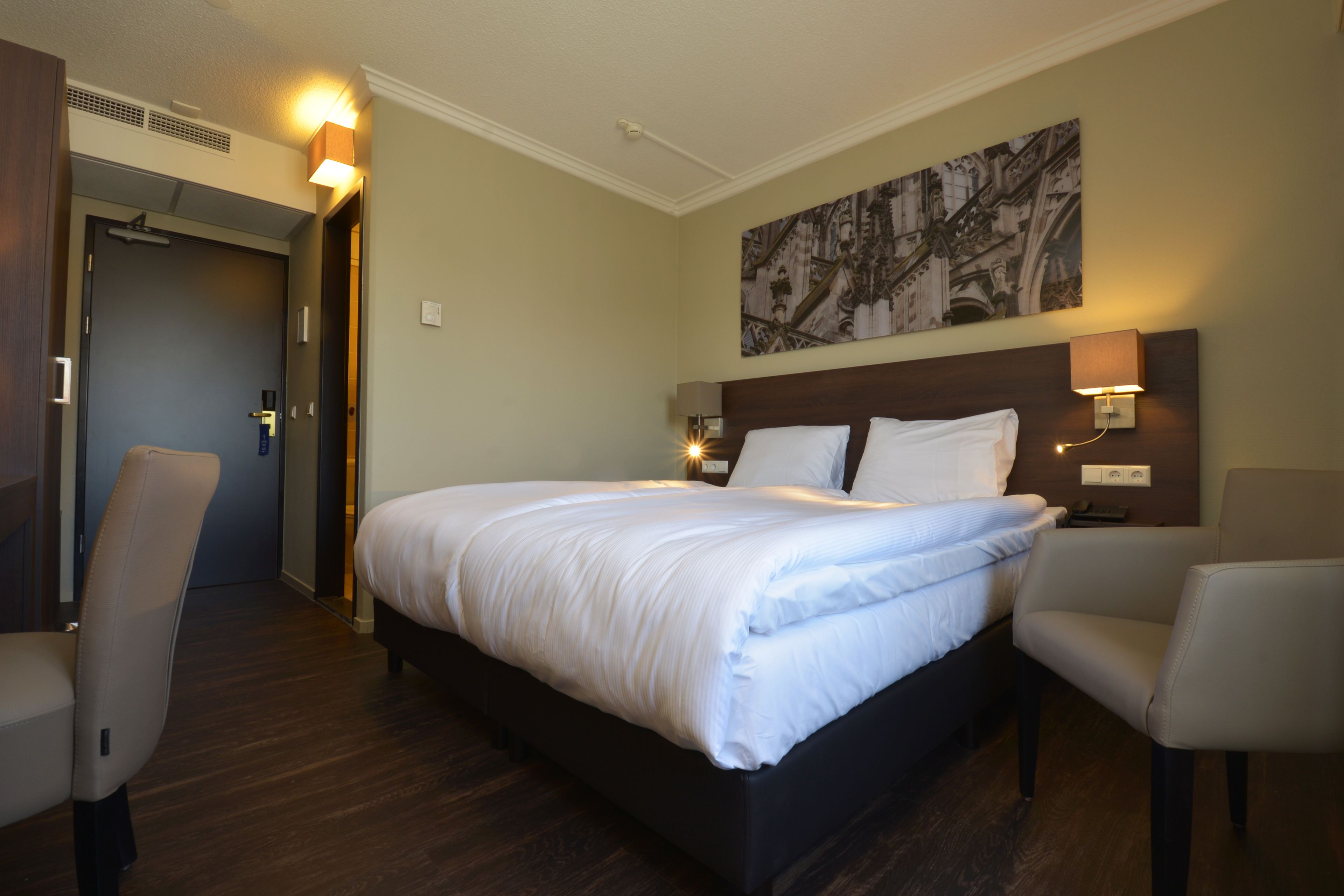 Quarto de hotel com uma cama grande, duas mesas de cabeceira, uma cadeira e um quadro na parede.