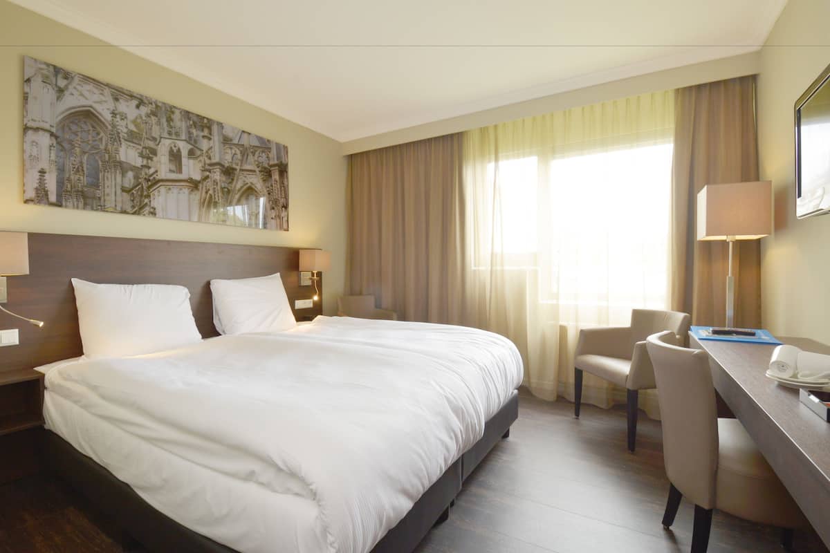 Quarto de hotel com uma cama grande, uma escrivaninha, uma cadeira, um abajur e um quadro na parede.