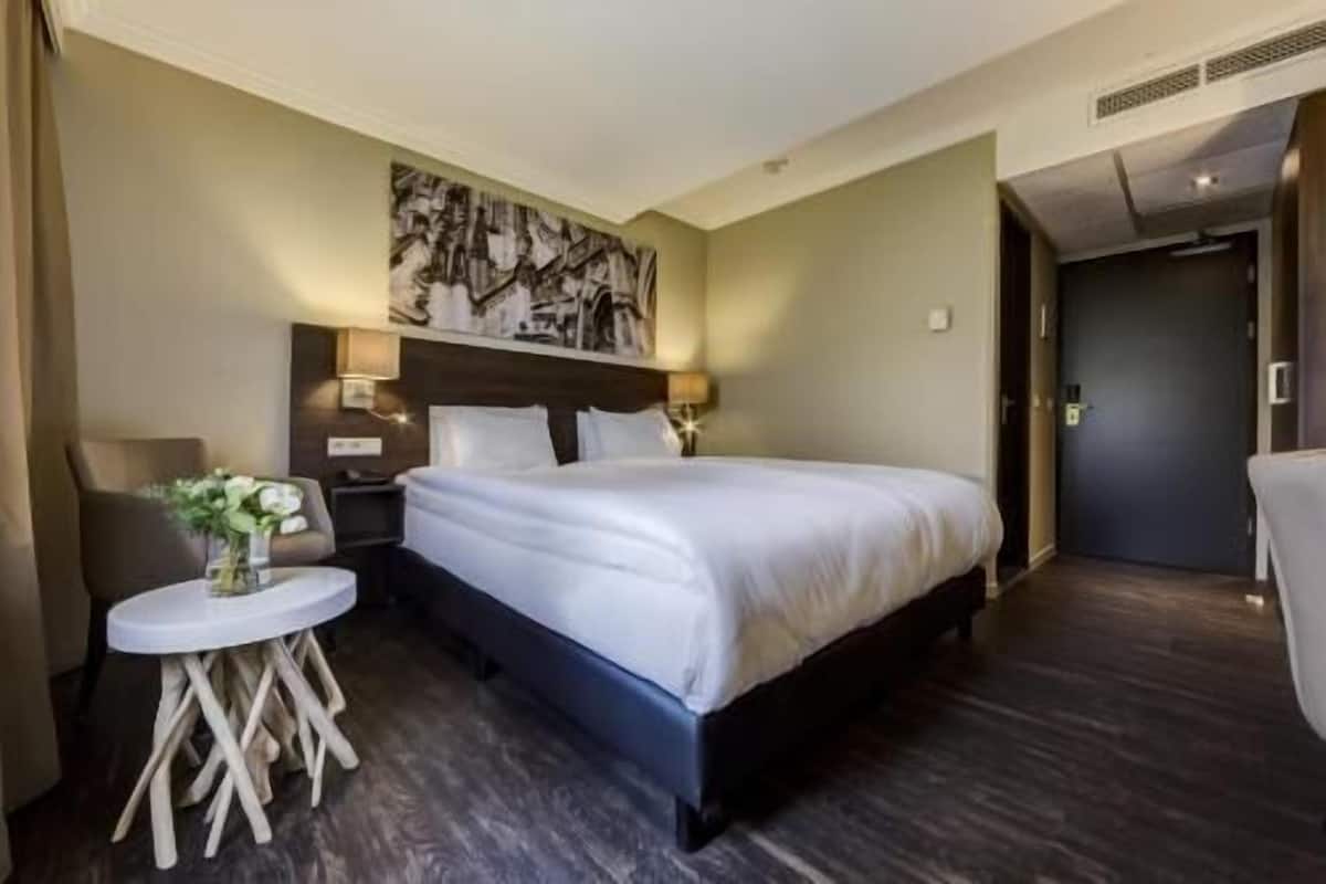 Quarto de hotel com cama, mesas de cabeceira, uma cadeira, uma pequena mesa redonda com um vaso e um grande quadro abstrato na parede.