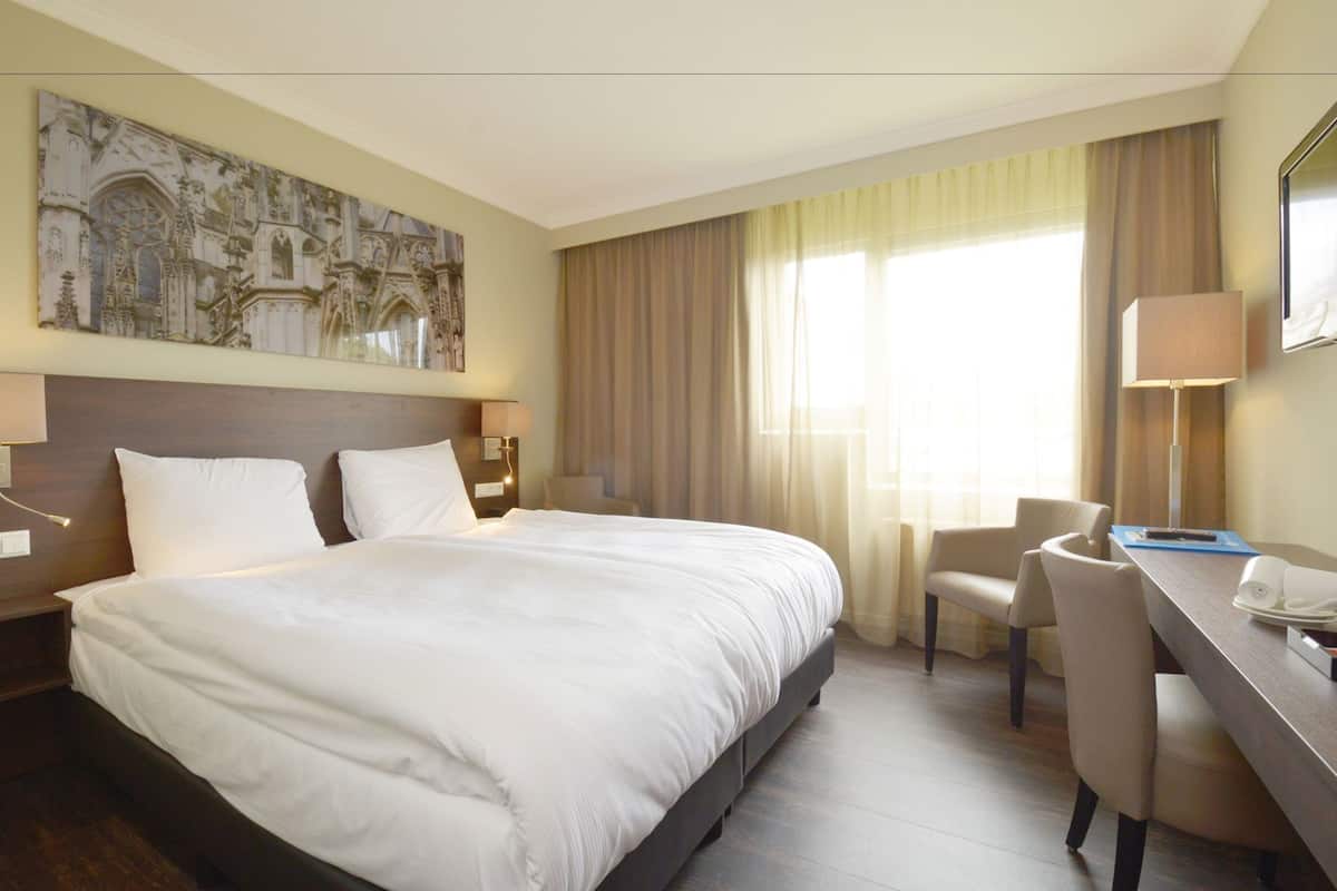 Quarto de hotel com uma cama grande, uma escrivaninha com cadeira, luminária de mesa e um quadro na parede.