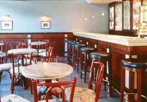 Uma área de bar com mesas de madeira com tampo de mármore e cadeiras vermelhas, banquetas de madeira e um balcão com luminária.