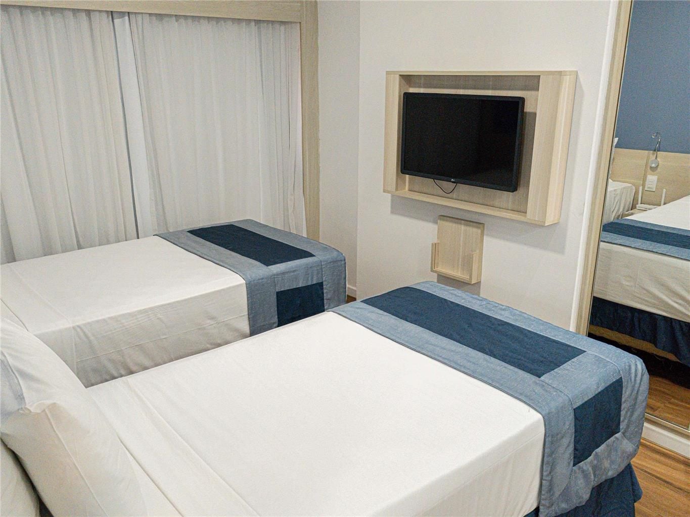 Quarto de hotel com duas camas, televisão fixada na parede e um espelho refletindo o ambiente.