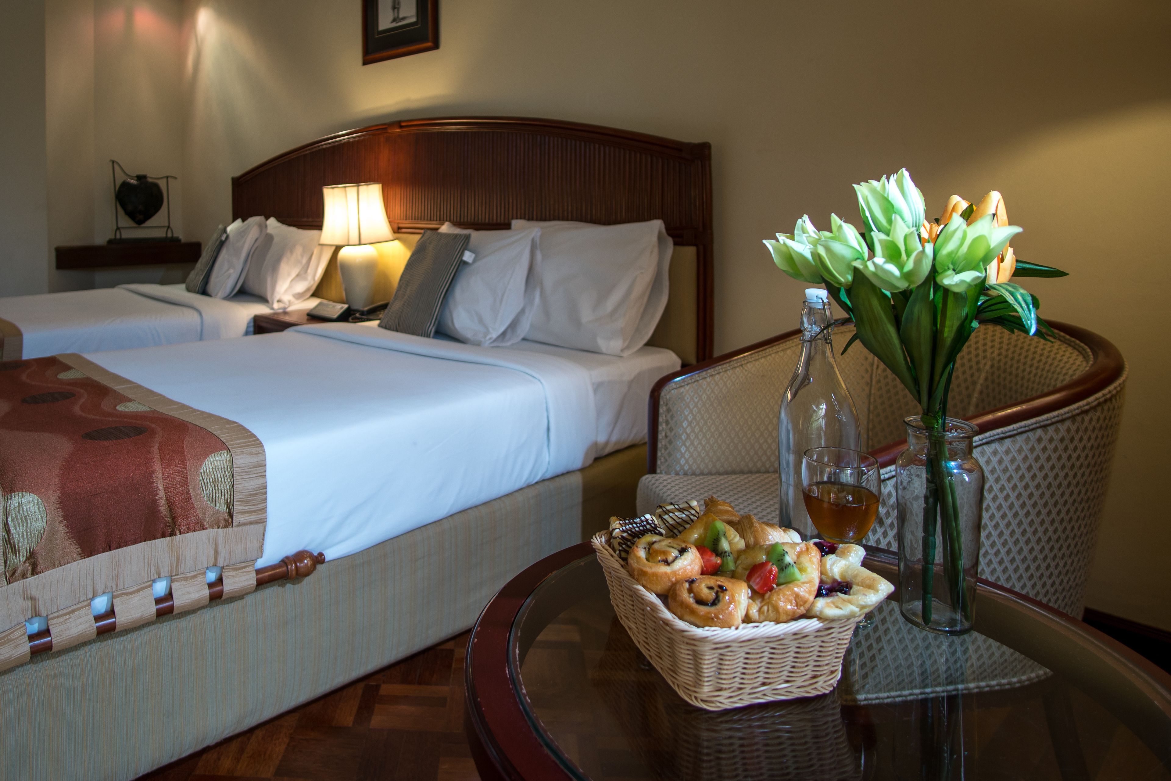 Quarto de hotel com duas camas, uma mesa de jantar com uma cesta de doces, um vaso com flores e uma garrafa de água.
