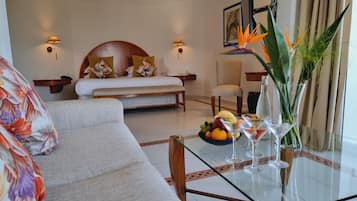 Quarto de hotel com uma cama, um sofá, uma mesa de jantar com frutas e copos de vinho, e uma planta decorativa.
