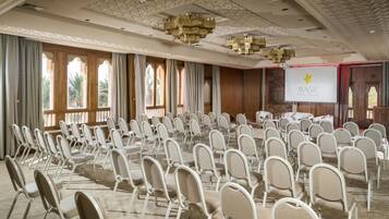 Uma sala de conferências com fileiras de cadeiras brancas, um palco com um banner da Magic Hotels & Resorts e detalhes decorativos no teto.