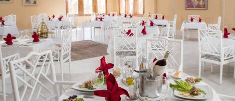 Uma sala de jantar formal com mesas e cadeiras brancas, dispostas para uma refeição.