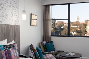 Quarto com uma janela grande, uma cama, duas cadeiras, uma mesa pequena e um quadro na parede.