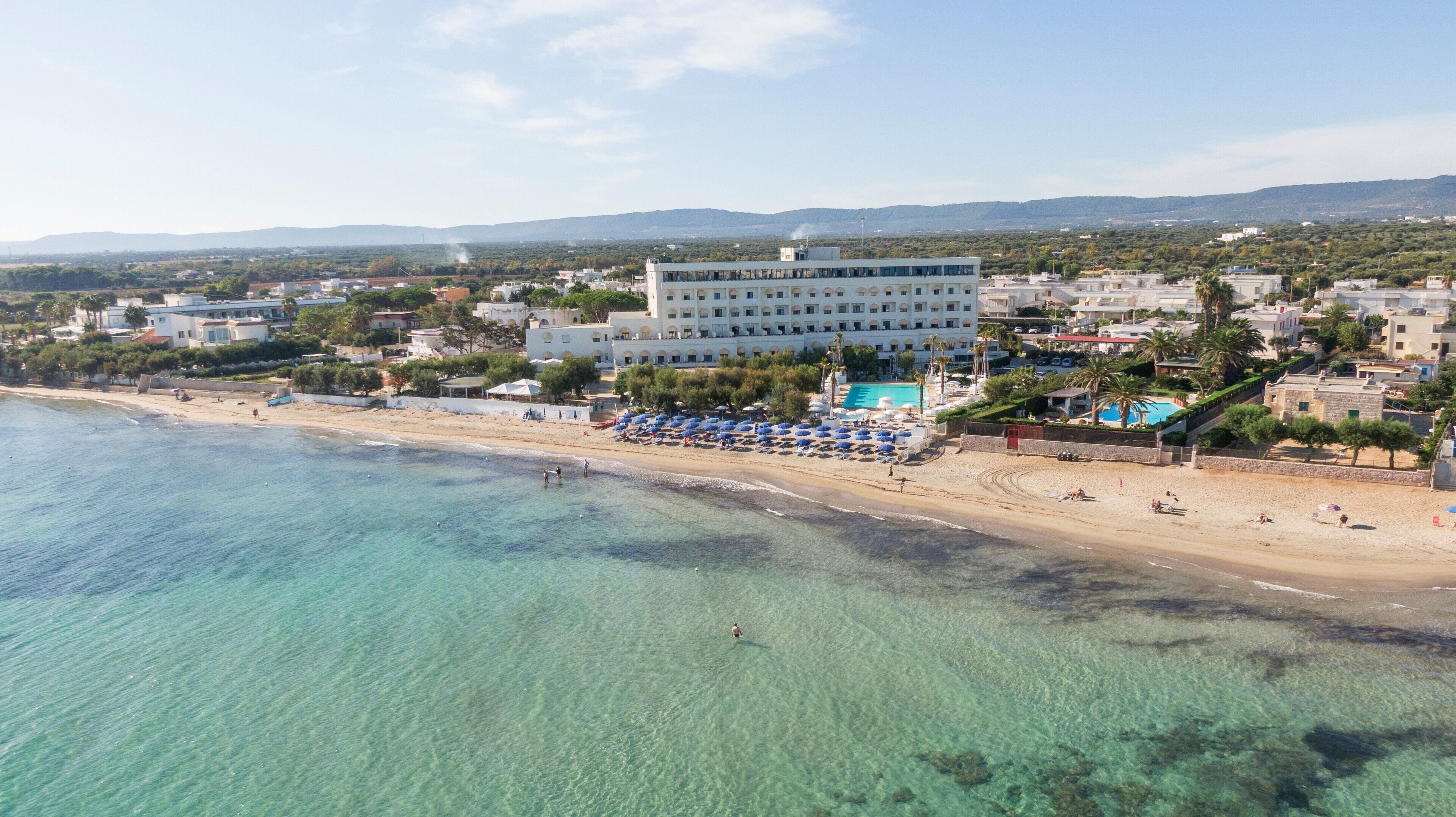 Um hotel à beira-mar, com praia, águas cristalinas e montanhas ao fundo.