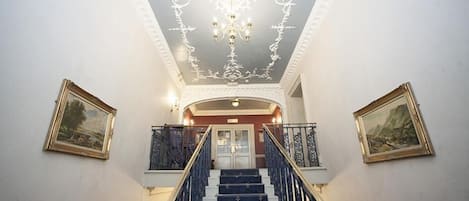 Uma escadaria imponente com corrimãos elaborados e um candelabro, levando a uma porta com moldura vermelha.