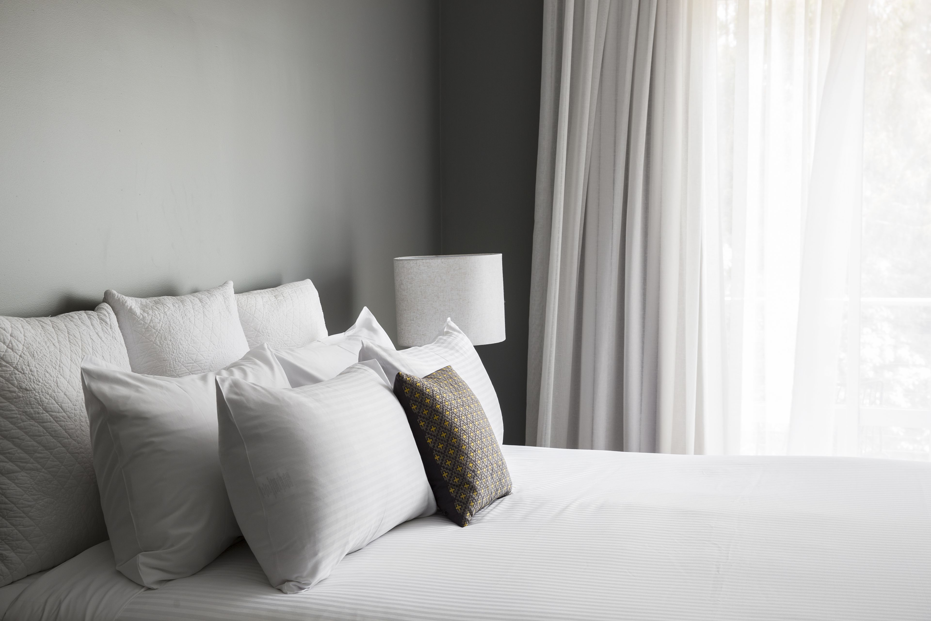 Cama bem arrumada com roupa de cama branca e várias almofadas, abajur e cortinas translúcidas que permitem a entrada de luz natural.
