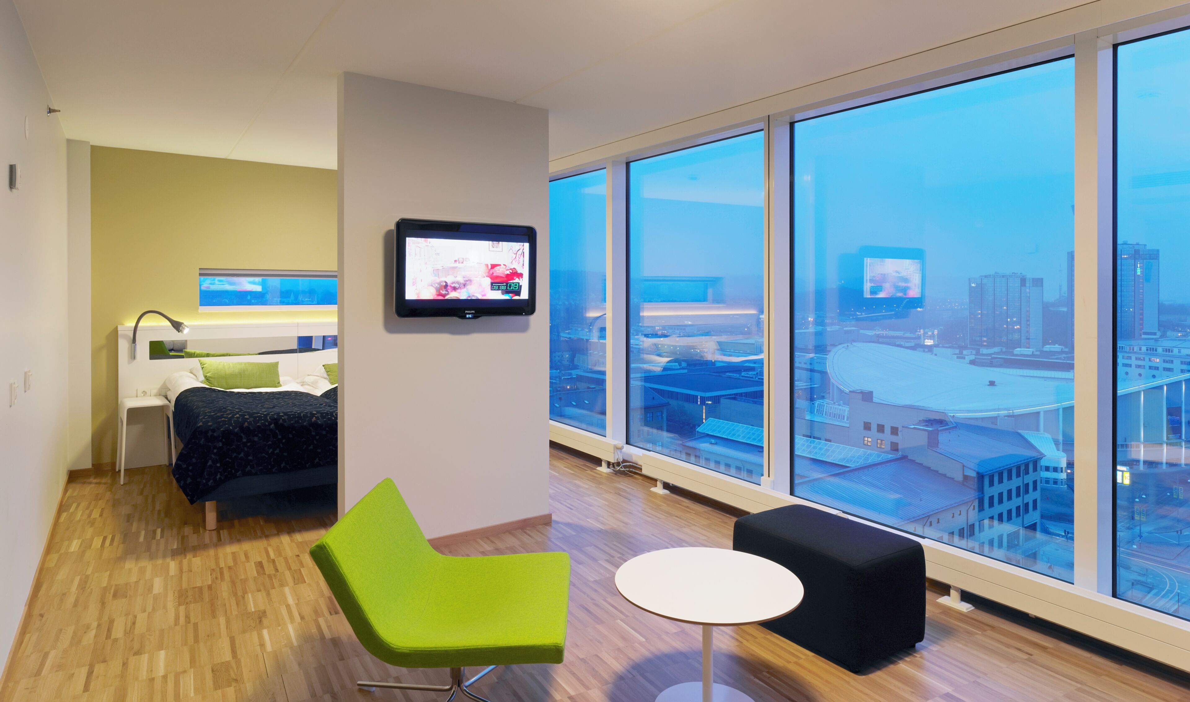 Quarto de hotel moderno com cama, televisão, cadeira verde, mesa pequena e amplas janelas com vista para a cidade.