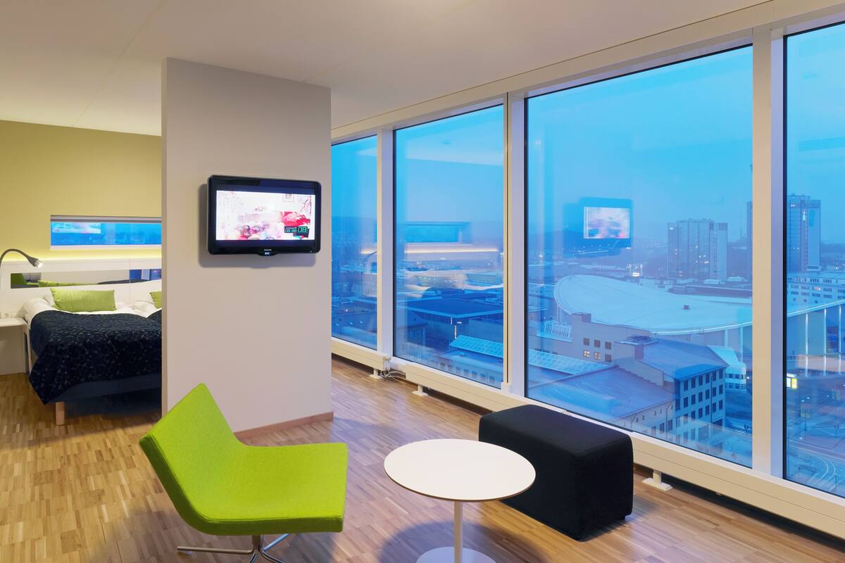 Quarto de hotel moderno com cama, televisão, cadeira verde, mesa pequena e amplas janelas com vista para a cidade.