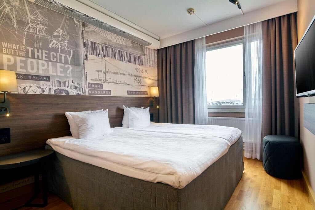 Quarto de hotel moderno com uma cama grande, papel de parede com tema urbano, televisão e uma janela com cortinas.