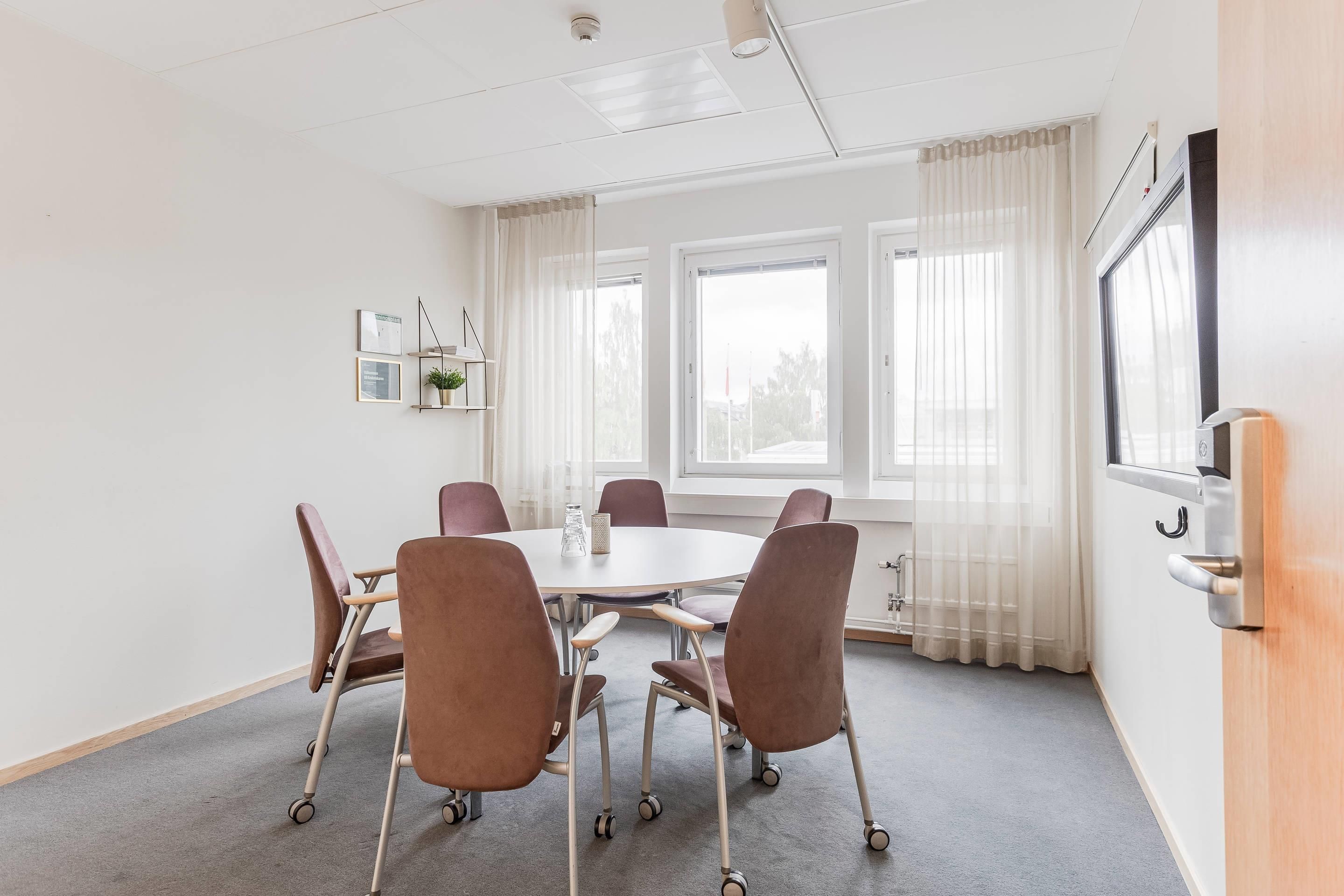 Uma sala de conferências com uma mesa redonda, seis cadeiras, um quadro branco e uma janela com cortinas.