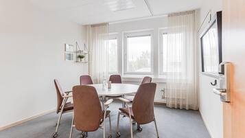 Uma sala de conferências com uma mesa redonda, seis cadeiras, um quadro branco e uma janela com cortinas.