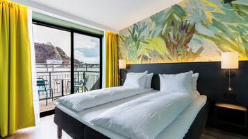 Quarto de hotel com cama, mesa de cabeceira, abajur, janela grande com cortinas amarelas e vista para uma varanda com cadeiras e corrimão.
