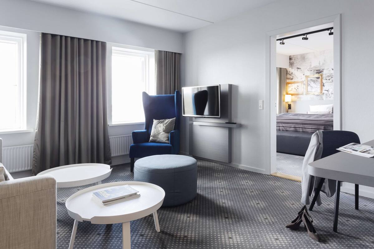 Quarto de hotel moderno com carpete cinza, poltrona azul, mesa redonda branca, banquinho cinza, TV de tela plana e cama com cabeceira cinza.