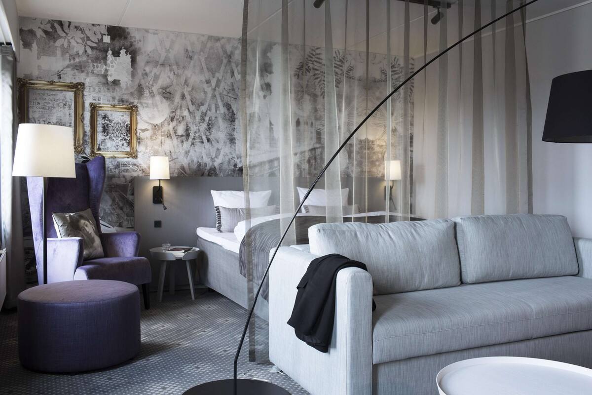 Quarto de hotel moderno com sofá cinzento, um banquinho redondo, uma pequena mesa lateral e uma cama com cabeceira branca.