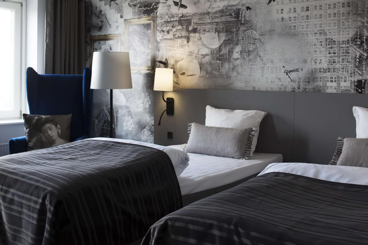 Quarto de hotel com duas camas, uma poltrona azul, um abajur e uma parede com design de mapa antigo.