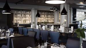 Um restaurante com mesas preparadas para refeições, uma estante de livros ao fundo e iluminação pendente.