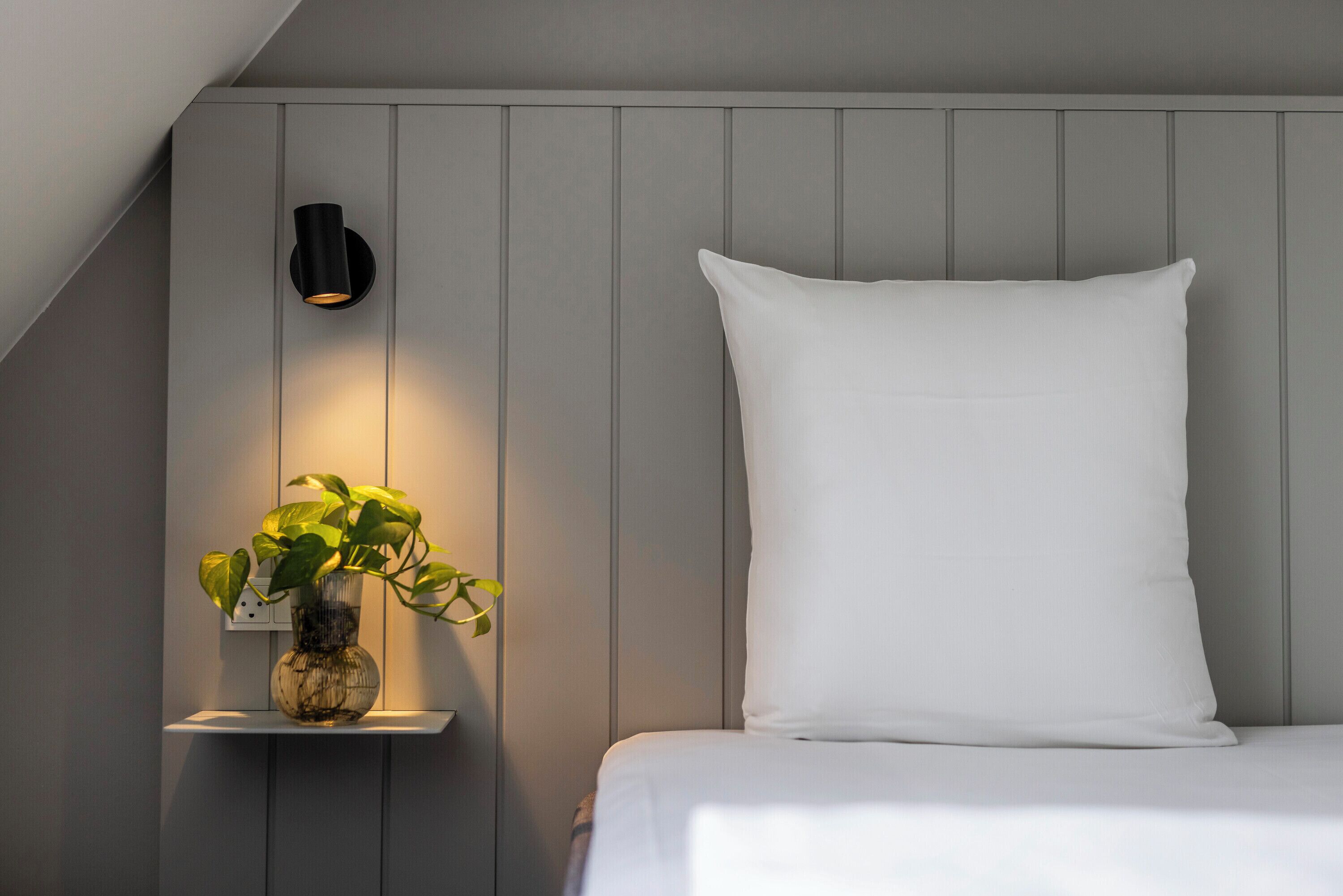 Quarto com cama, planta na mesa de cabeceira, luminária na parede e prateleira.
