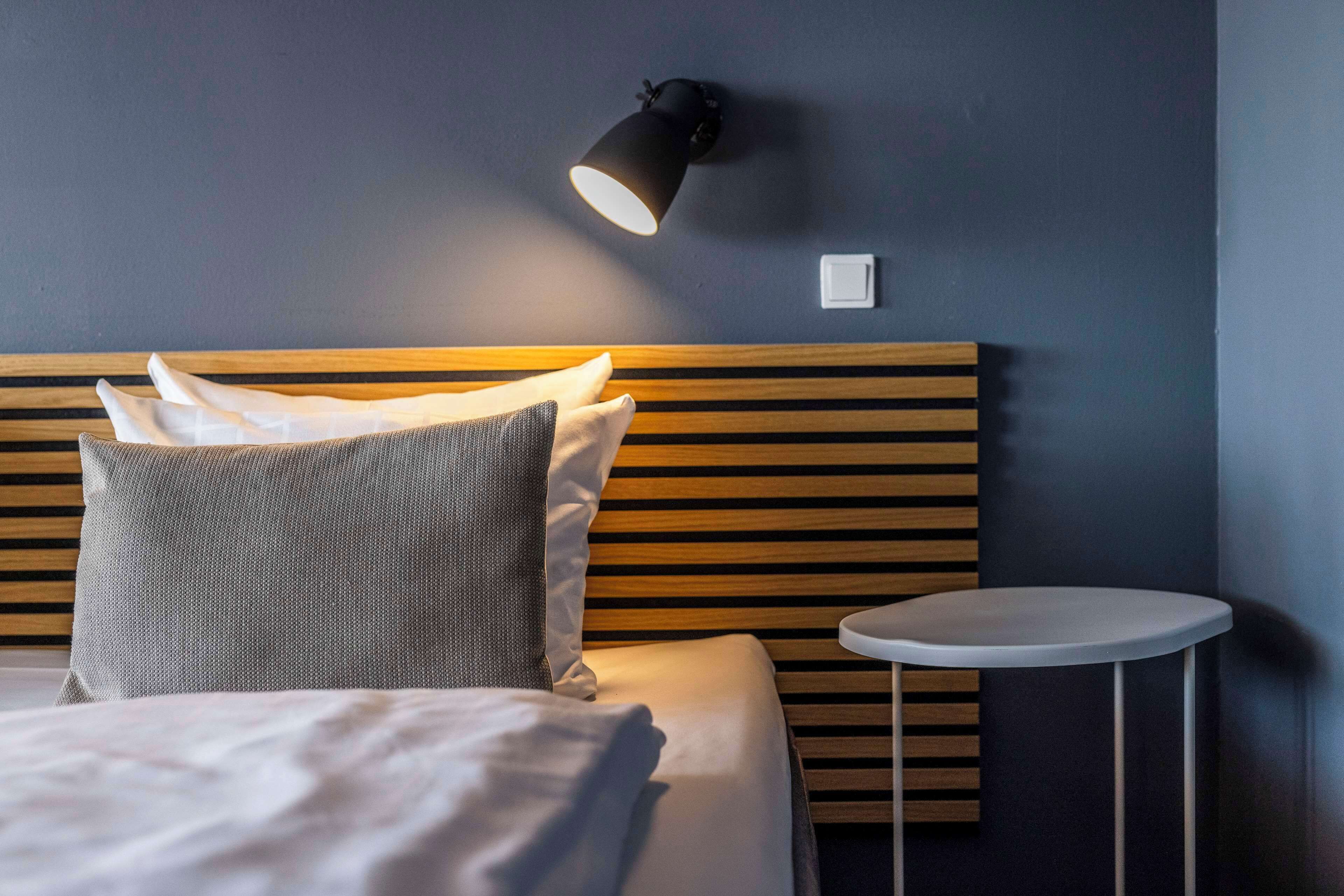 Uma cama bem arrumada com lençóis brancos e duas almofadas, uma mesa de cabeceira redonda e branca, e uma luminária fixada na parede.