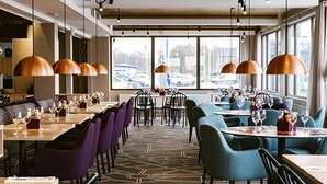 Um restaurante com várias mesas e cadeiras, grandes janelas e luminárias pendentes.