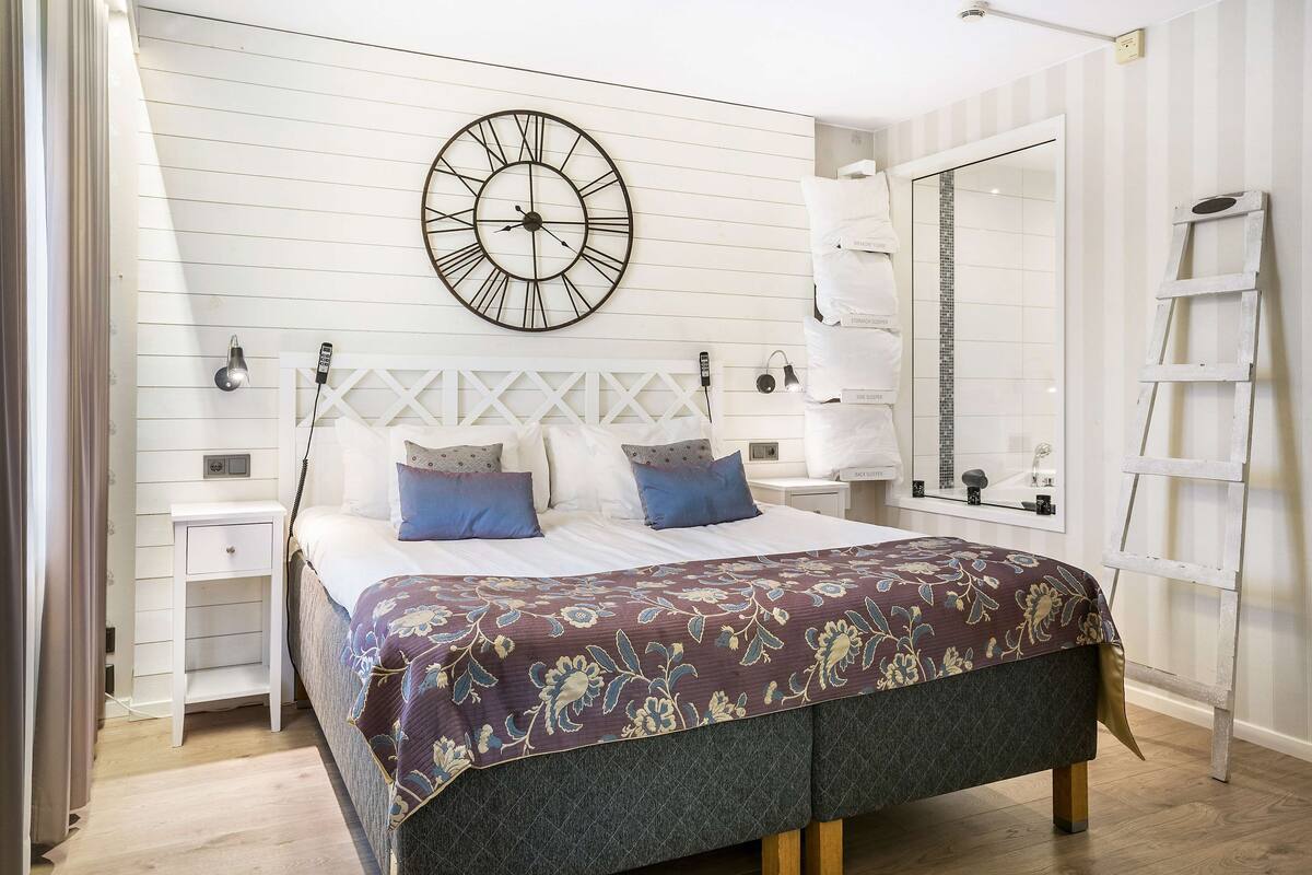 Quarto bem arrumado com uma cama grande, um relógio de parede, um espelho e uma escada.
