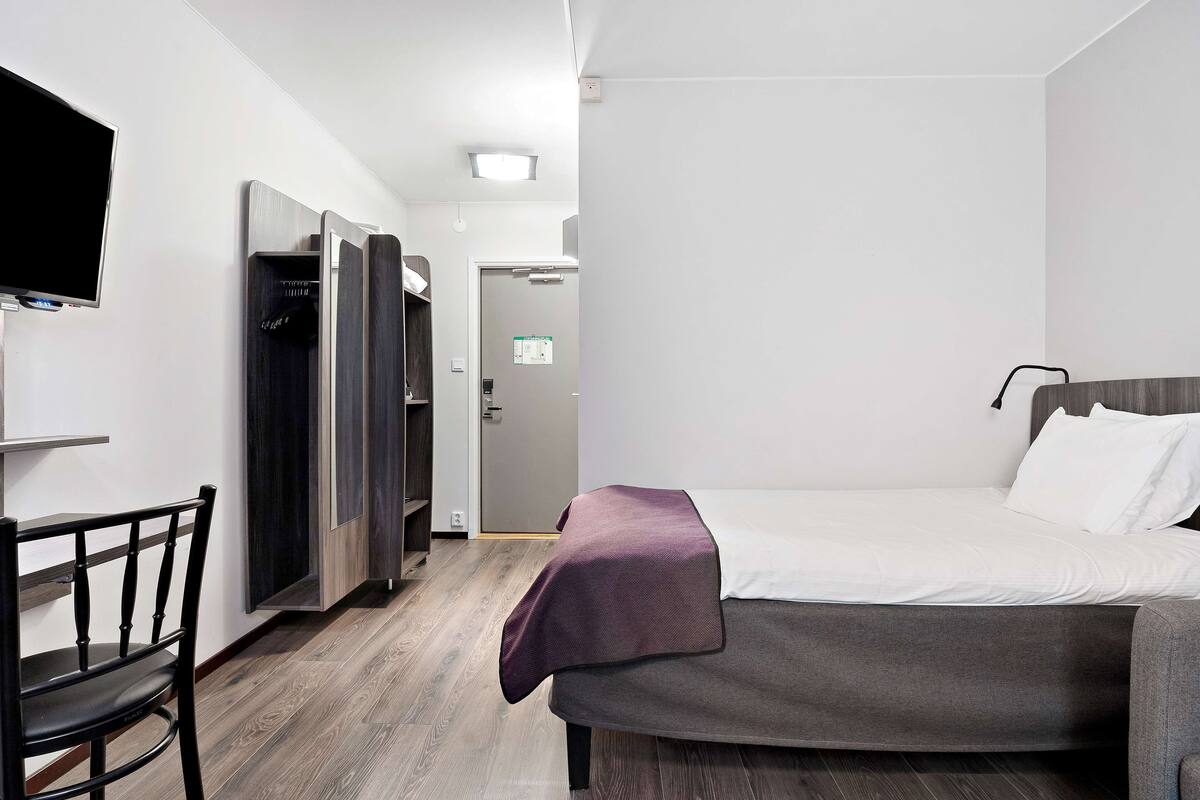 Quarto de hotel com cama, escrivaninha, cadeira, televisão e guarda-roupa.