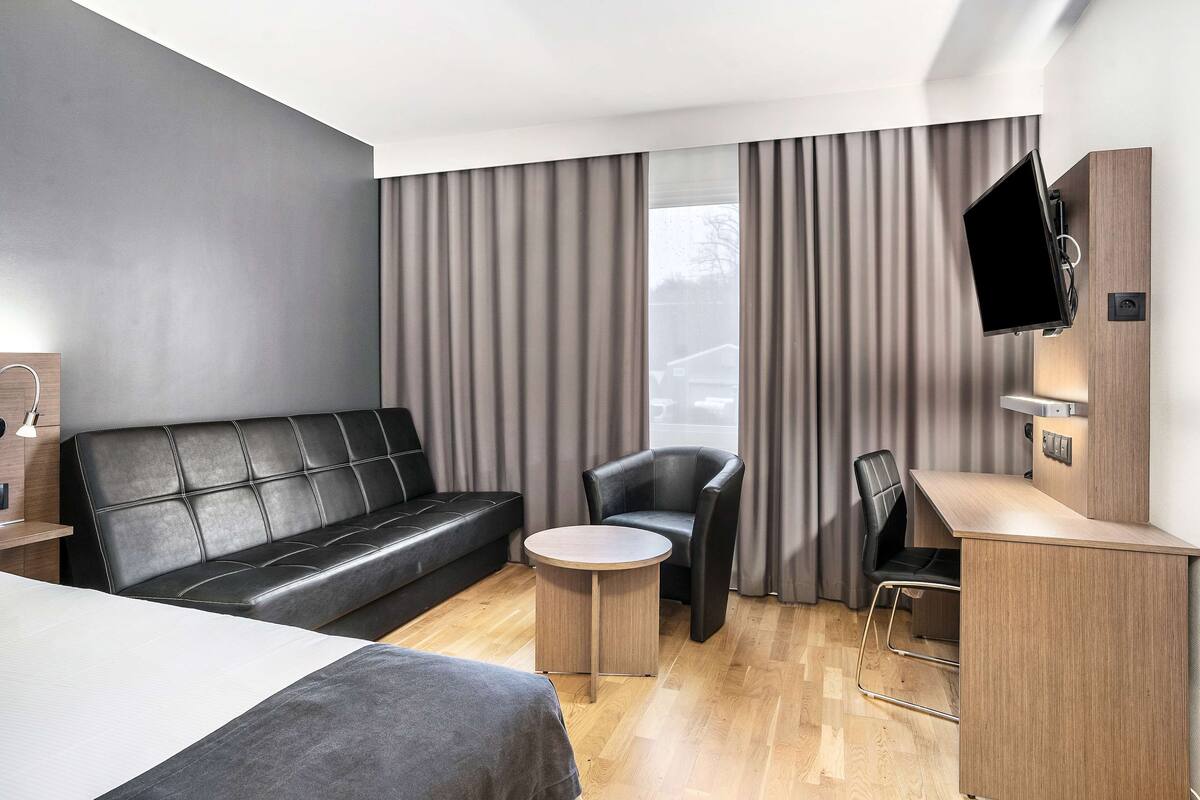 Quarto de hotel moderno com sofá de couro, mesa redonda pequena, escrivaninha com cadeira e TV fixada na parede.