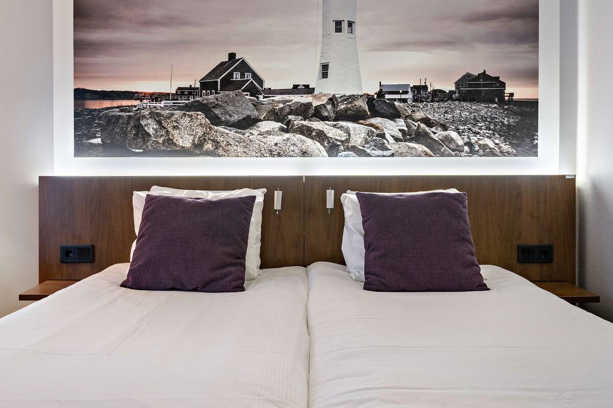 Uma cama com roupa de cama branca e duas almofadas, cabeceira de madeira e uma grande moldura com uma imagem costeira de um farol.